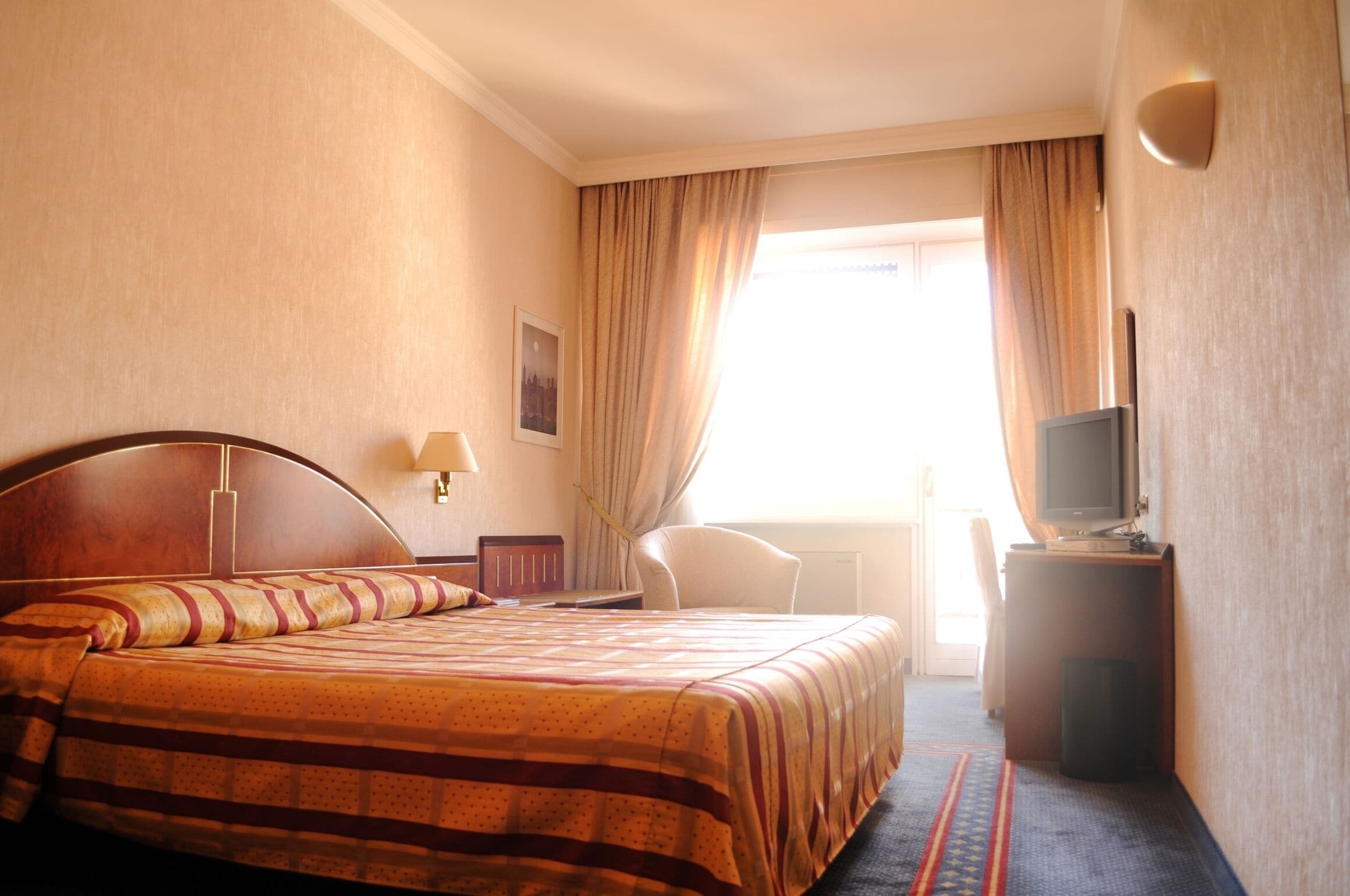 Quarto de hotel com uma cama grande, uma escrivaninha com televisão, uma cadeira e uma janela com cortinas.