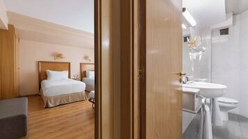 Quarto de hotel com chão de madeira, cama com roupa de cama branca, mesa de cabeceira com abajur e banheiro com pia e vaso sanitário.