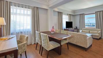 Quarto de hotel com mesa de jantar, cadeiras, sofá, televisão e vista para o porto.