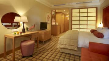 Quarto de hotel com uma cama, uma secretária com computador, uma cadeira, uma pequena mesa com fruta e um espelho.