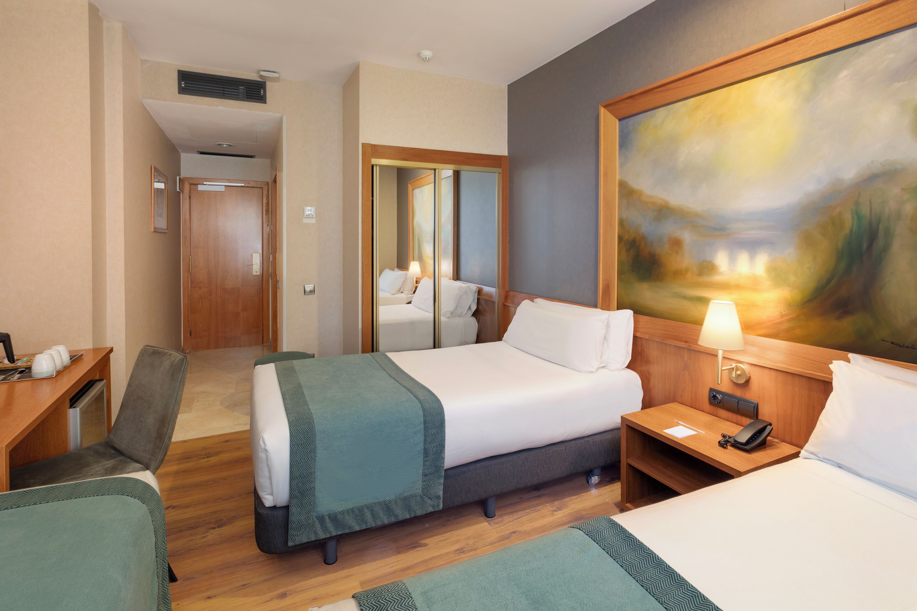 Quarto de hotel com cama, escrivaninha, cadeira, mesa de cabeceira, luminária e um quadro na parede.