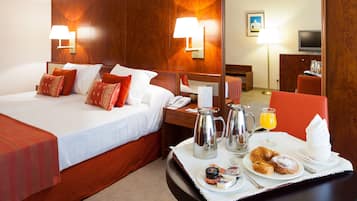 Quarto de hotel com cama, mesa de cabeceira e bandeja de café da manhã com pães, café e suco.