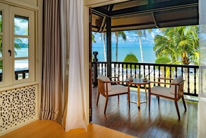 Um deck de madeira com duas cadeiras e uma mesa, com vista para uma praia com palmeiras e o oceano.
