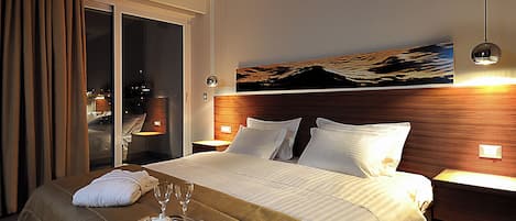 Quarto de hotel com uma cama grande, cabeceira de madeira, um quadro sobre a cama e um tabuleiro com toalha, taça de vinho e chá na cama.