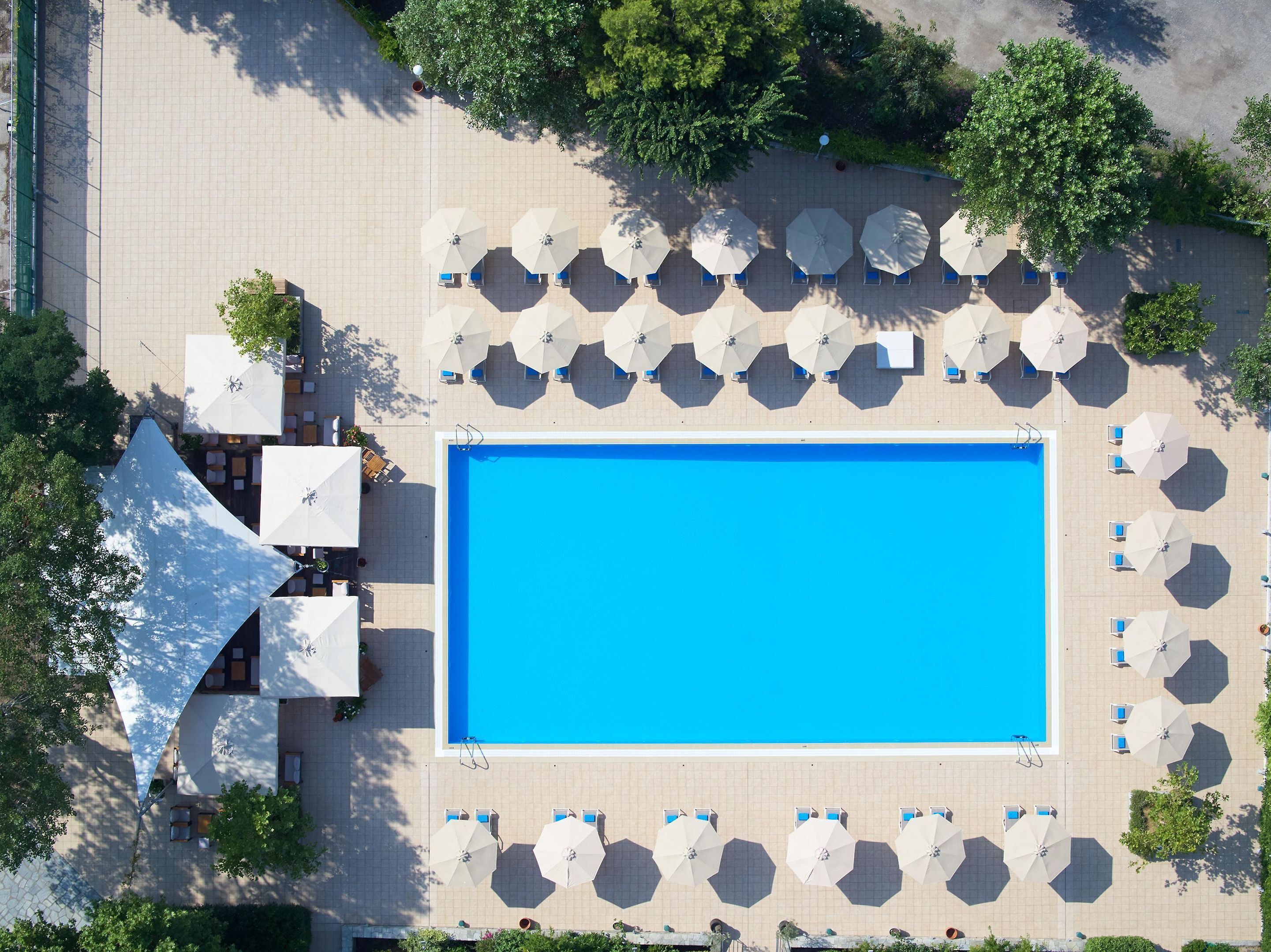 Vista aérea de uma área de piscina com vários espreguiçadeiras e guarda-sóis, cercada por árvores e um caminho de azulejos.