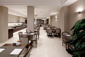 Uma área de refeições de hotel com várias mesas e cadeiras, um balcão de buffet e uma grande planta em vaso.