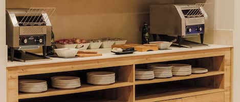 Espaço do buffet de café da manhã com variedade de pães, doces e uma máquina de café. Há prateleiras de madeira, um canto para café e uma área de estar com cadeiras.