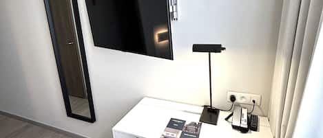 Quarto de hotel com escrivaninha branca, televisão de tela plana fixada na parede, luminária de mesa e uma pequena lixeira.