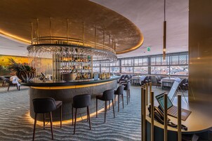 Uma área de bar moderna com balcão curvo, vários bancos e vista da cidade por grandes janelas.
