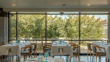 Restaurante com mesas preparadas para refeições, janelas amplas com vista para árvores e edifícios.