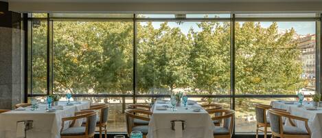 Restaurante com mesas preparadas para refeições, janelas amplas com vista para árvores e edifícios.
