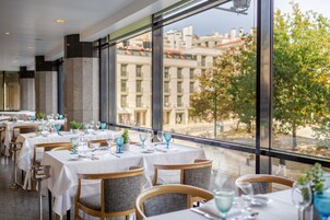 Um restaurante com várias mesas preparadas para refeições, grandes janelas com vista para edifícios e árvores.