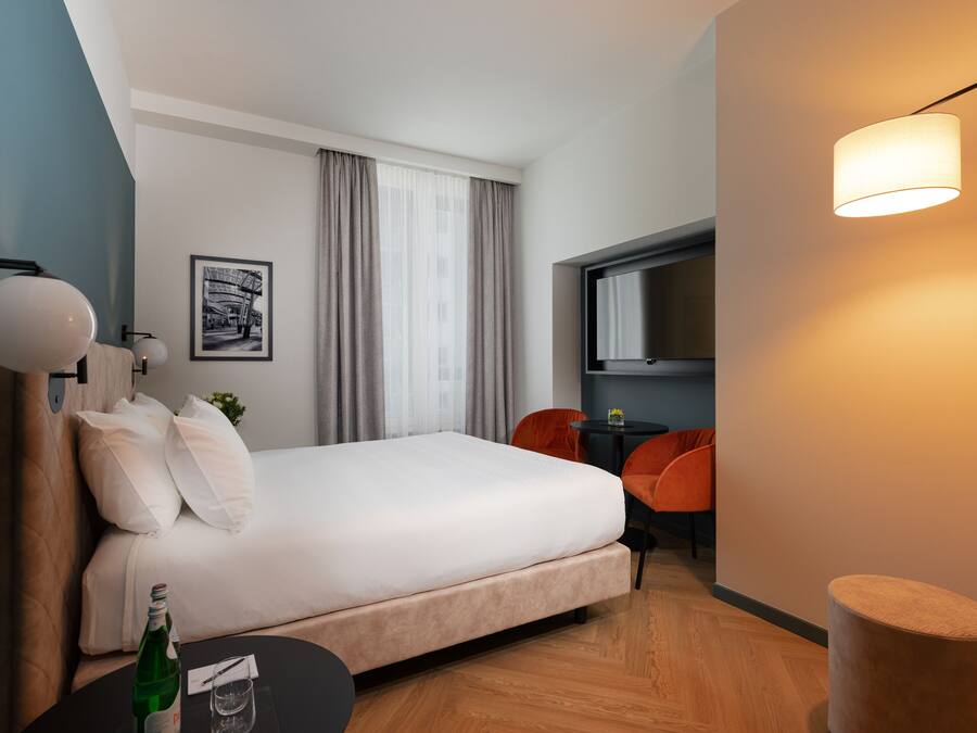 Quarto de hotel moderno com cama, escrivaninha, cadeira, abajur e televisão.