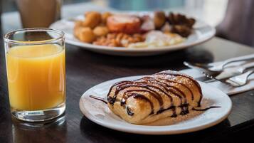 Mesa de café da manhã com copo de suco de laranja, prato de croissants e prato com variedade de doces.
