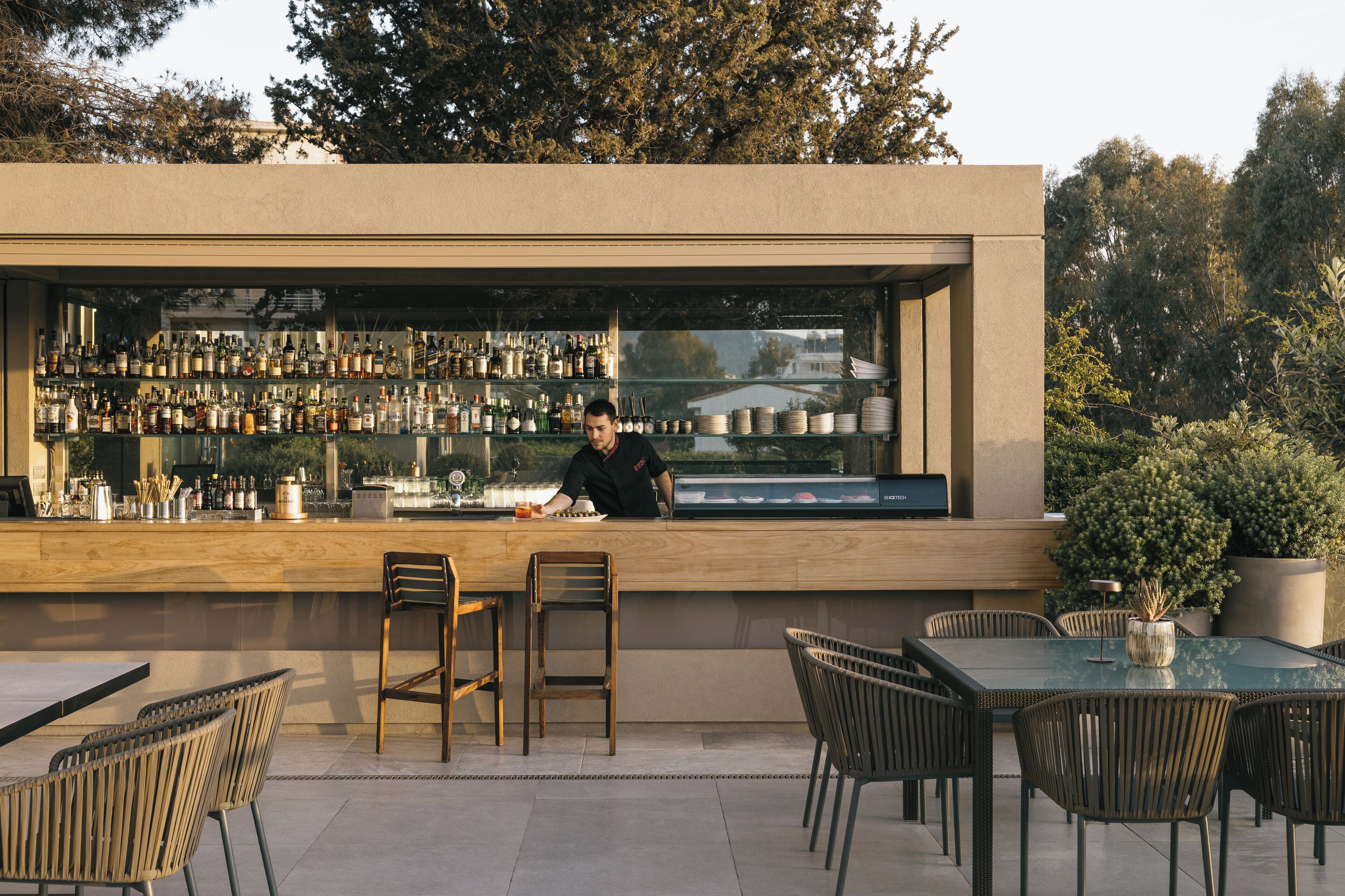 Uma área de bar ao ar livre com um bartender, cadeiras de madeira e uma mesa de vidro.