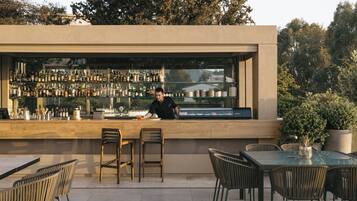 Uma área de bar ao ar livre com um bartender, cadeiras de madeira e uma mesa de vidro.