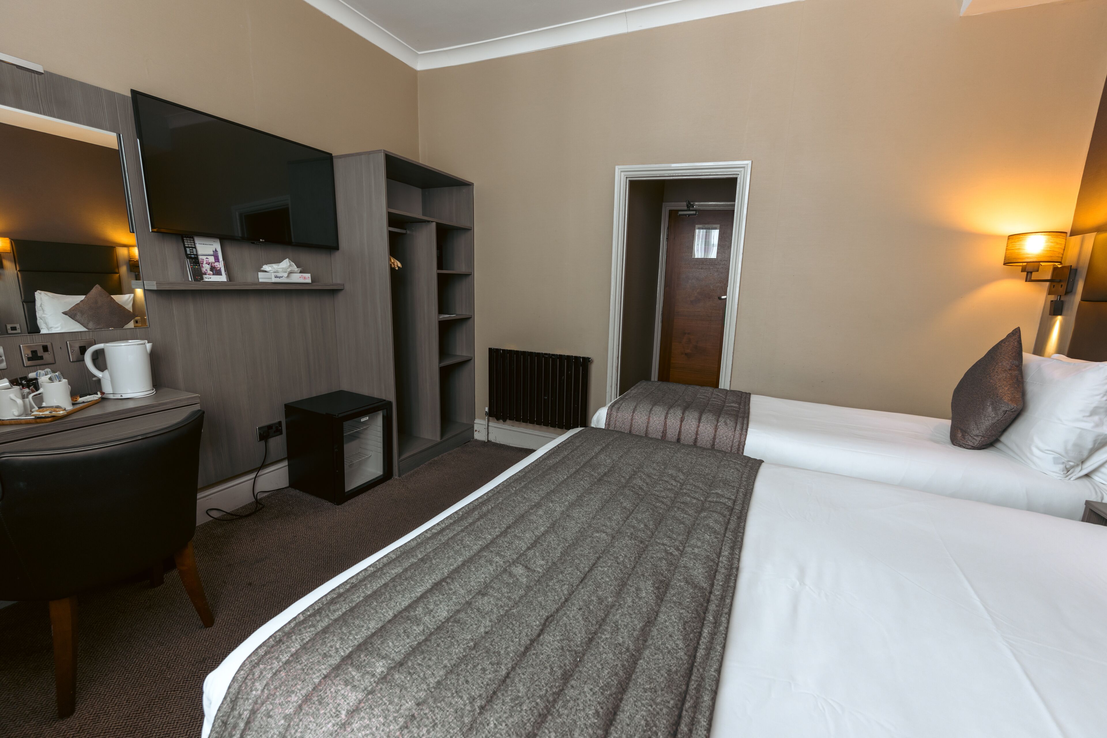 Quarto de hotel com cama, escrivaninha com chaleira, televisão e guarda-roupa.