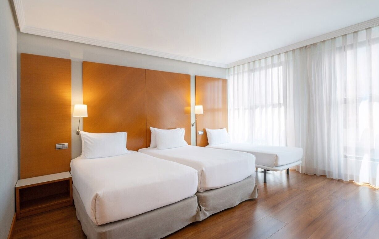 Quarto de hotel com duas camas, um espaço de estar pequeno e uma janela grande com cortinas leves.