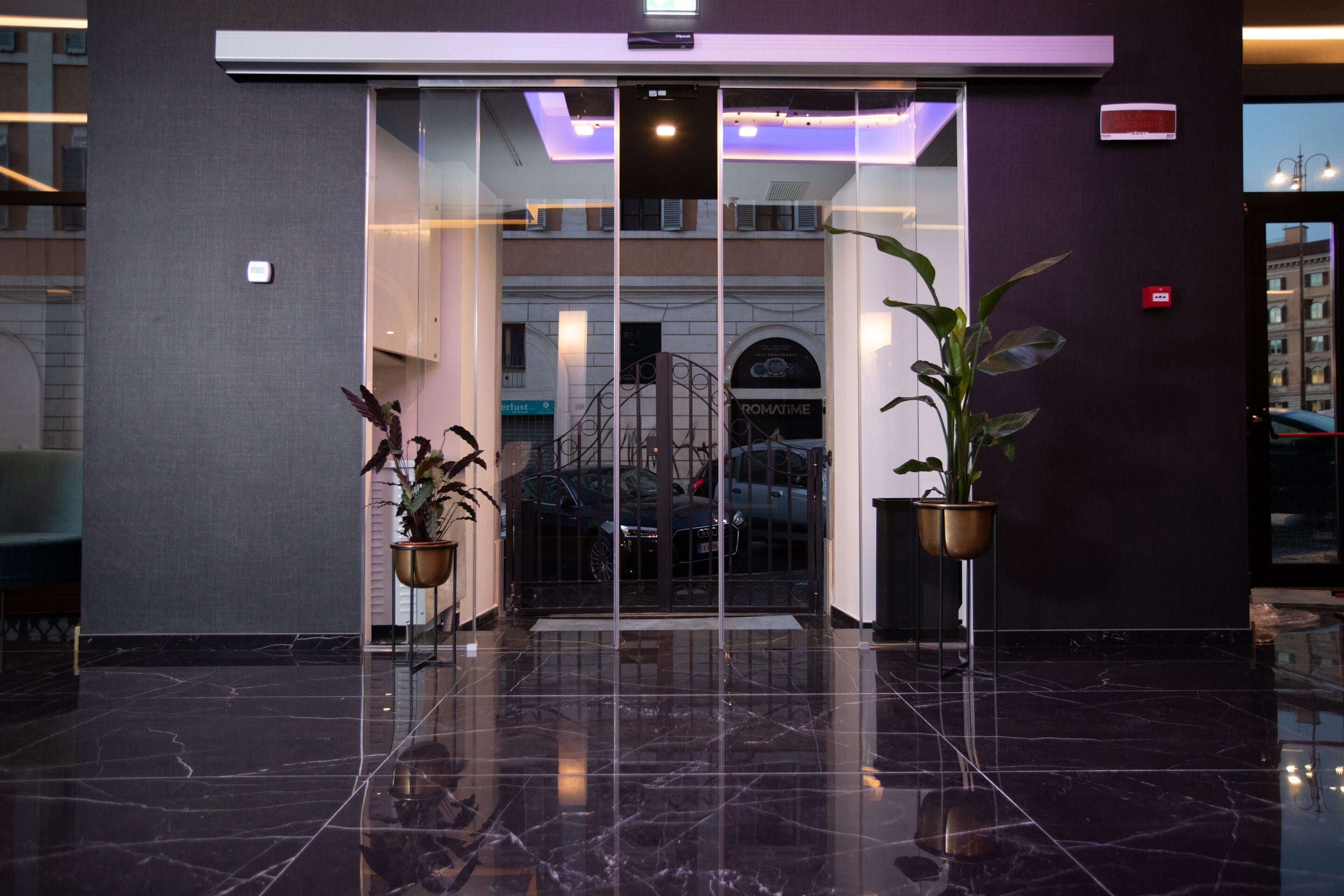 Entrada de um hotel moderno, com porta de vidro, plantas em vasos e chão de mármore.