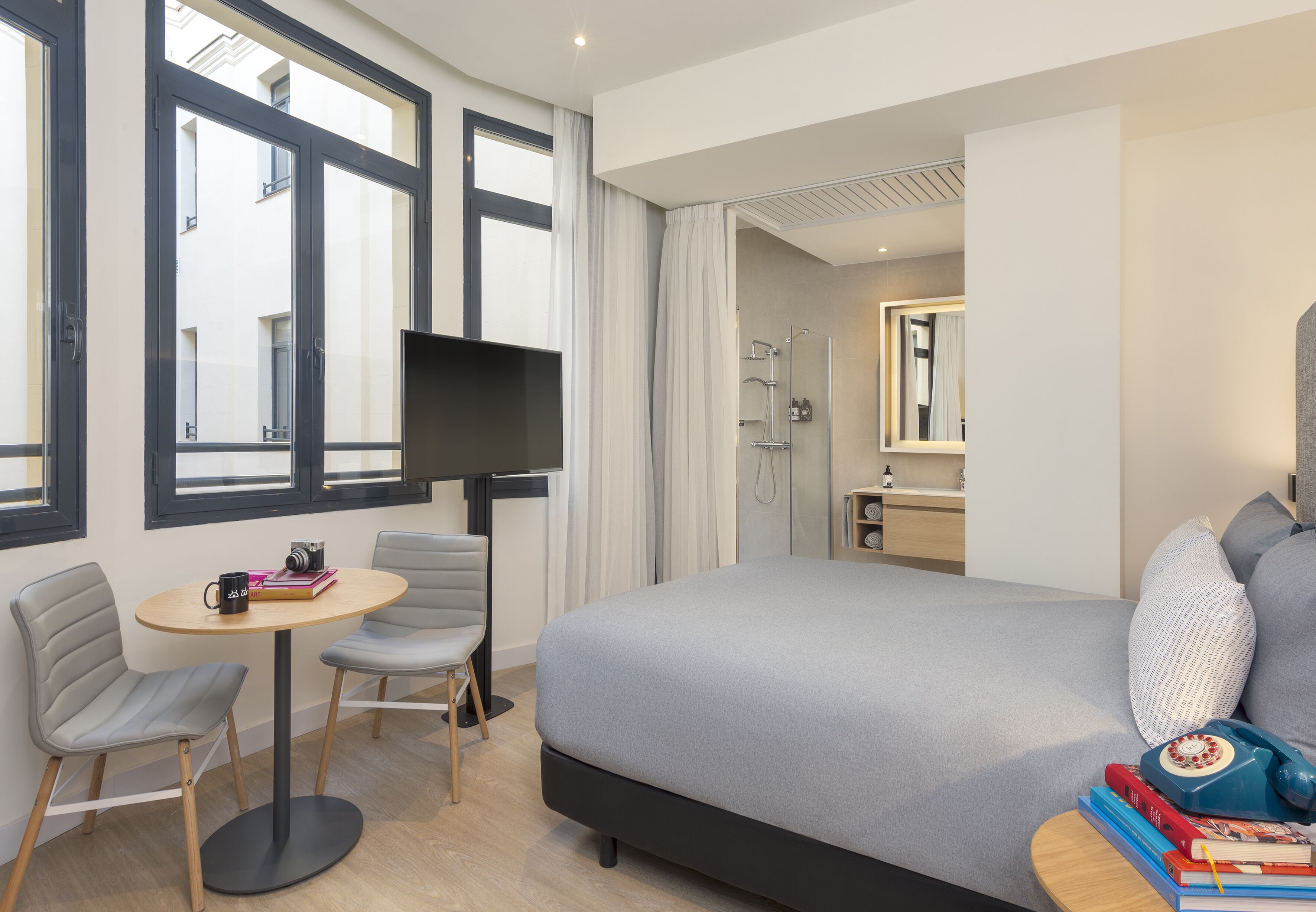 Quarto de hotel moderno com uma cama grande, uma televisão, uma pequena mesa com cadeiras e um banheiro visível através de uma porta aberta.