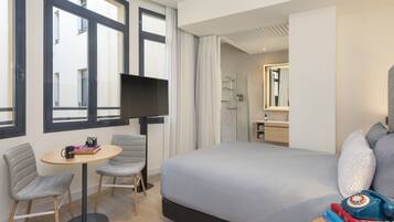 Quarto de hotel moderno com uma cama grande, uma televisão, uma pequena mesa com cadeiras e um banheiro visível através de uma porta aberta.