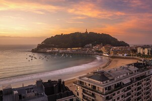 Paisagem costeira com uma colina imponente, uma praia e diversos edifícios.