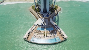 Vista aérea de uma ilha resort, com piscina central, diversas áreas de estar e um edifício alto.