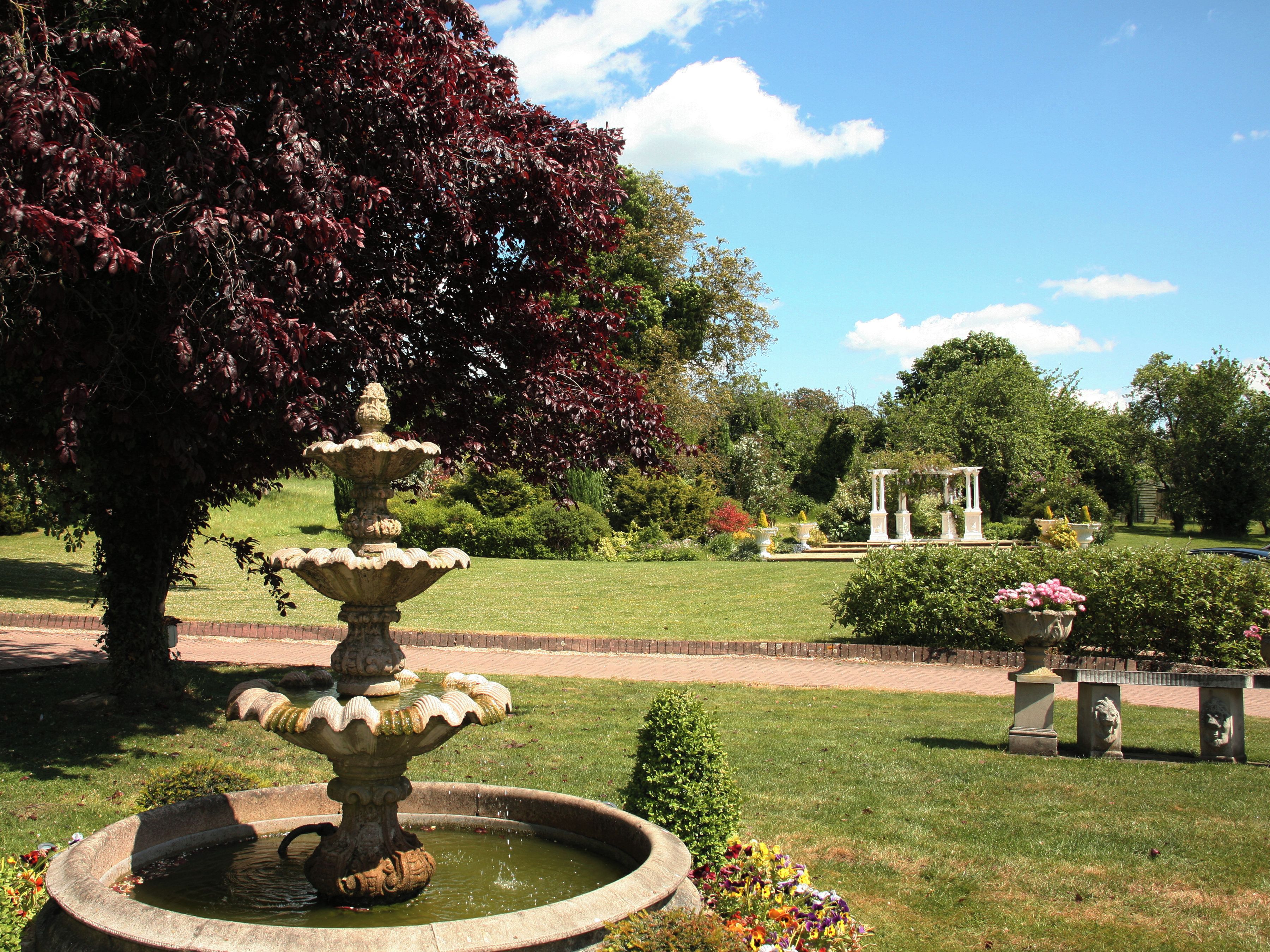 Um jardim com uma fonte em vários níveis, um coreto e diversos arbustos e flores.