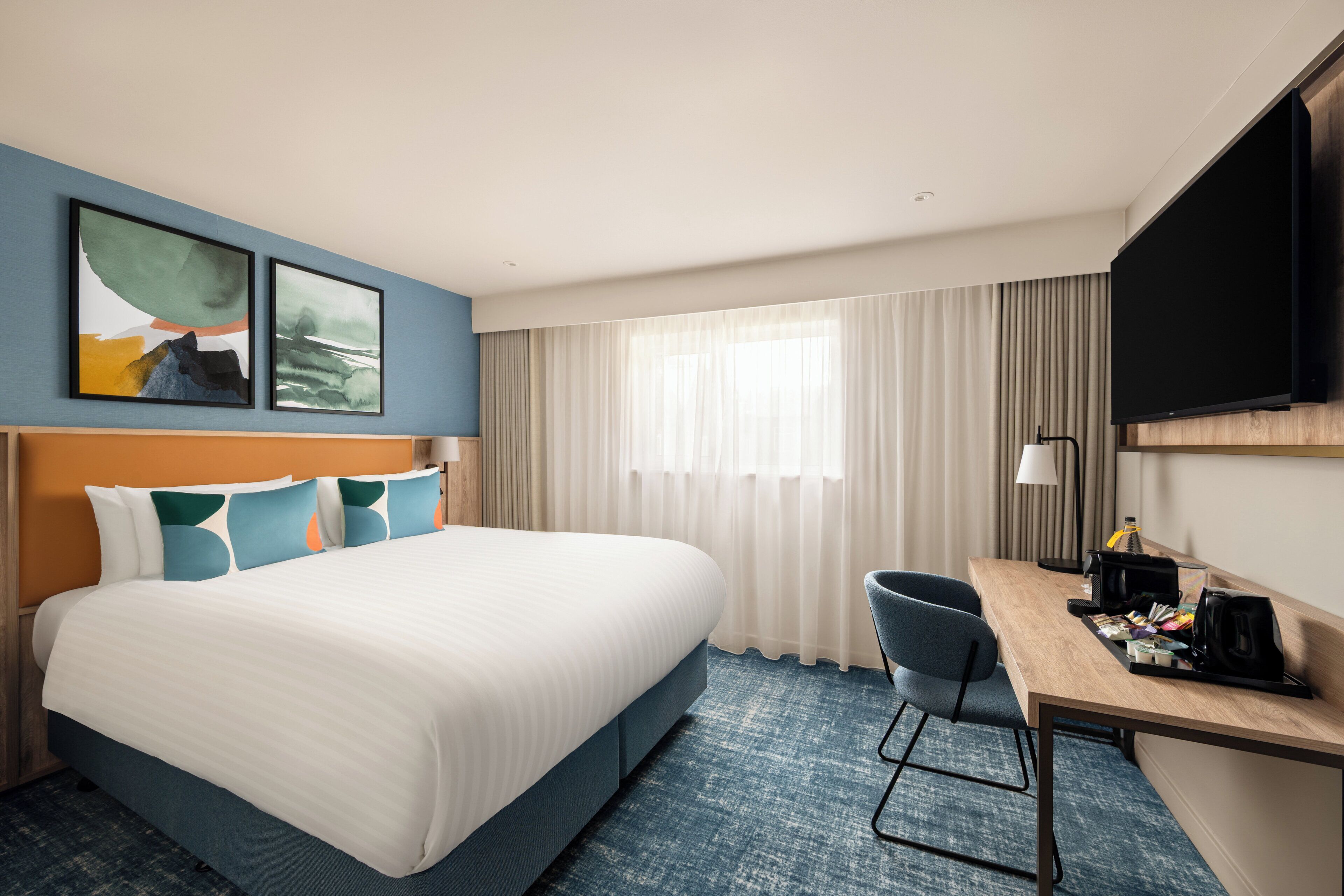 Quarto de hotel com uma cama grande, uma escrivaninha, uma cadeira, televisão de tela plana e obras de arte na parede.
