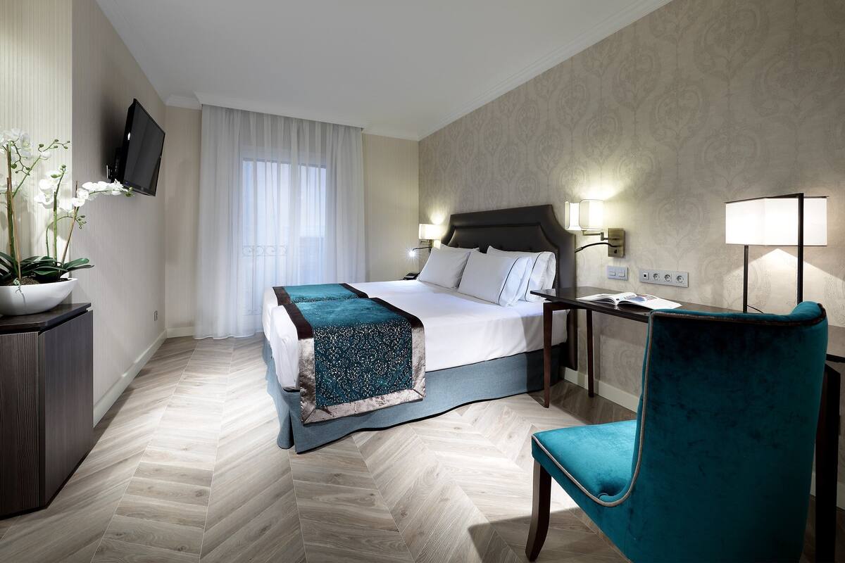 Quarto de hotel com cama, mesas de cabeceira, televisão na parede, planta em vaso e janela com cortinas.