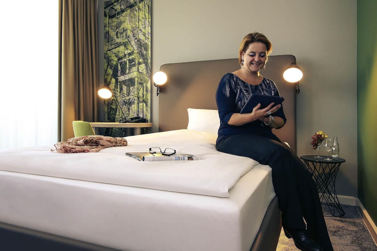Quarto de hotel com cama, mesa de cabeceira com abajur, uma obra de arte na parede e uma mesinha com um vaso e copos.