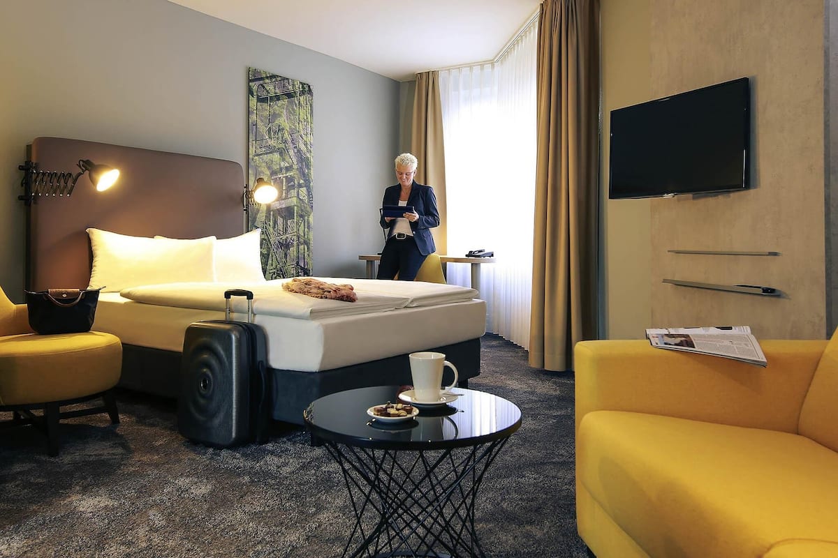 Quarto de hotel com cama, escrivaninha, cadeira, televisão e janela com cortinas.