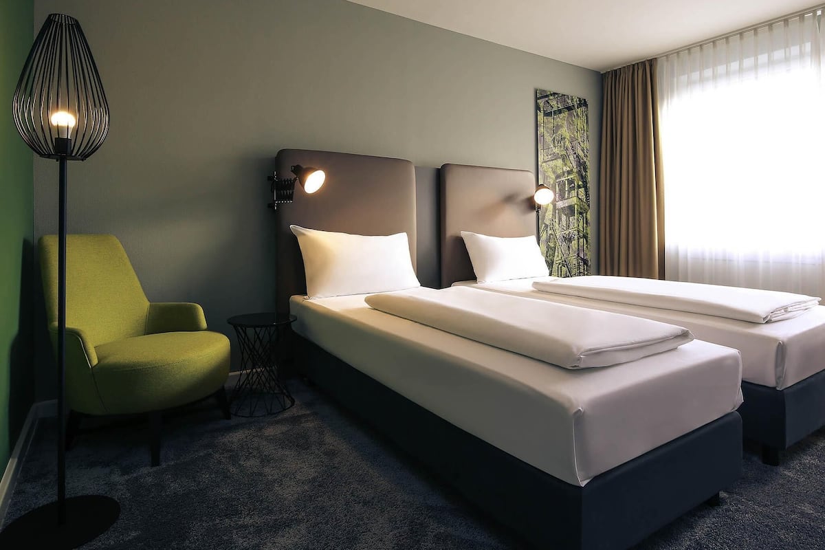 Quarto de hotel com duas camas, uma poltrona verde, um abajur e uma janela com cortinas.