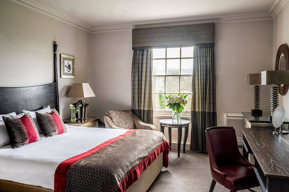 Quarto de hotel com uma cama grande, uma escrivaninha, uma cadeira, uma mesinha de centro com um vaso de flores e uma janela com cortinas.