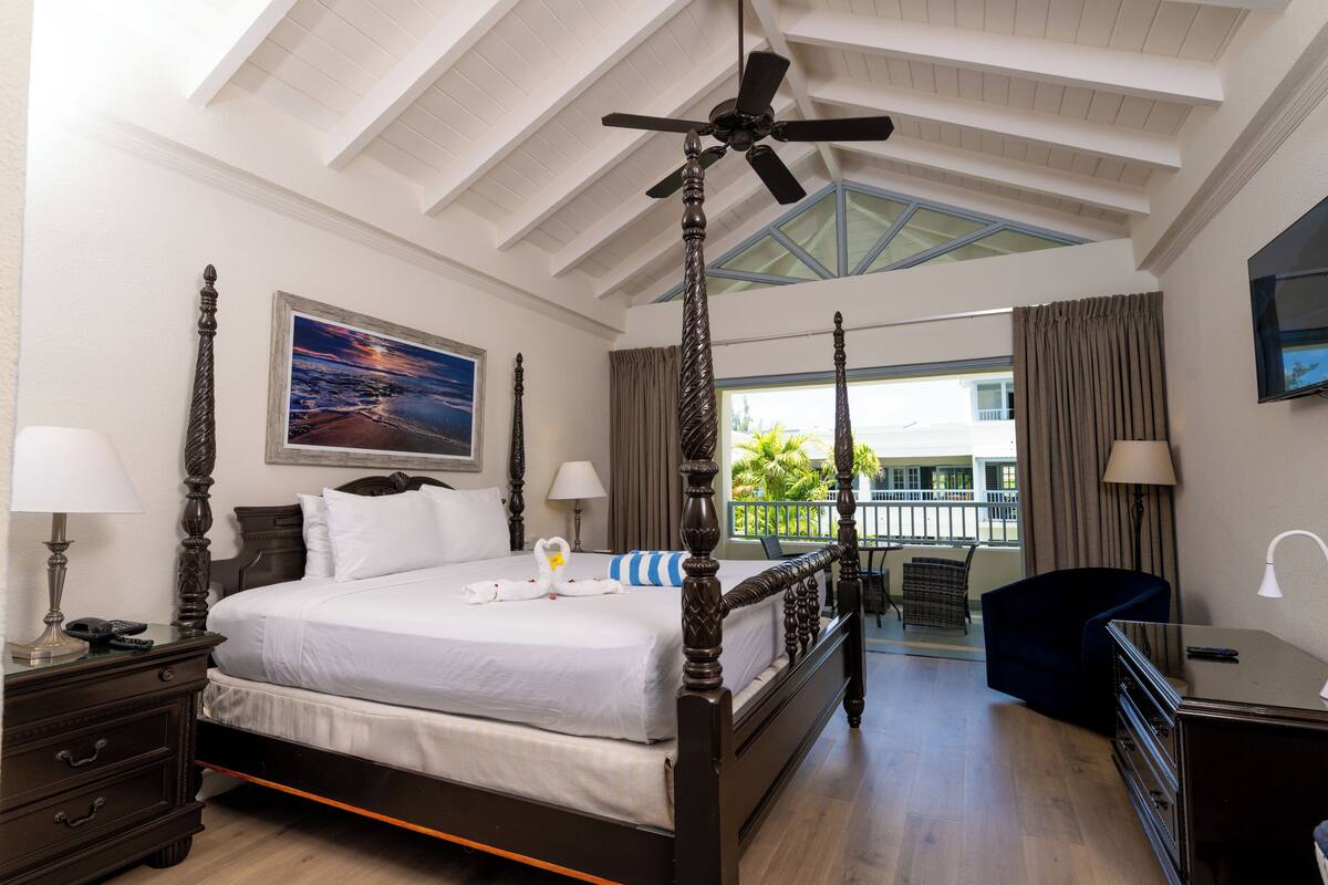 Uma cama de dossel com um ventilador de teto, um quadro na parede, uma varanda com vista e uma escrivaninha com abajur.