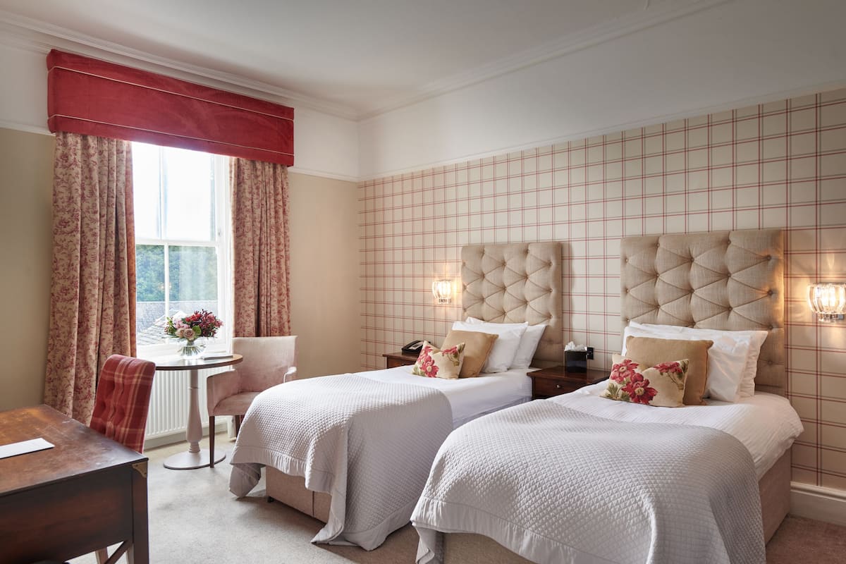 Quarto de hotel com duas camas, uma escrivaninha, uma cadeira e uma janela com cortinas.