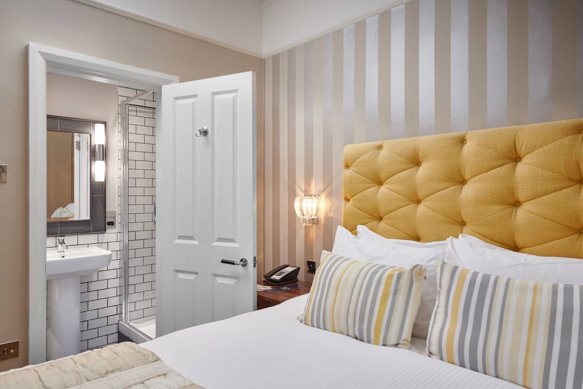 Quarto de hotel com cama, mesa de cabeceira, telefone, espelho e banheiro com pia e chuveiro.
