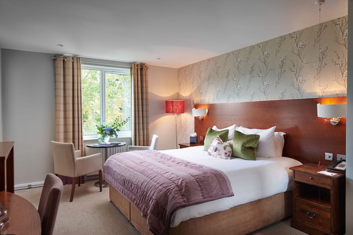 Quarto de hotel com uma cama grande, uma escrivaninha, uma cadeira, uma mesinha com um vaso de flores e uma janela com cortinas.