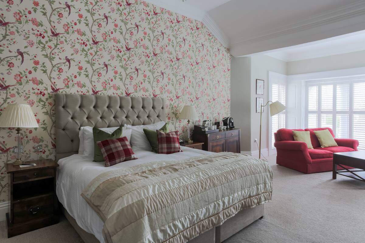 Quarto com cabeceira estofada, cama grande com várias almofadas, mesa de cabeceira com abajur, poltrona vermelha e janela com persianas.