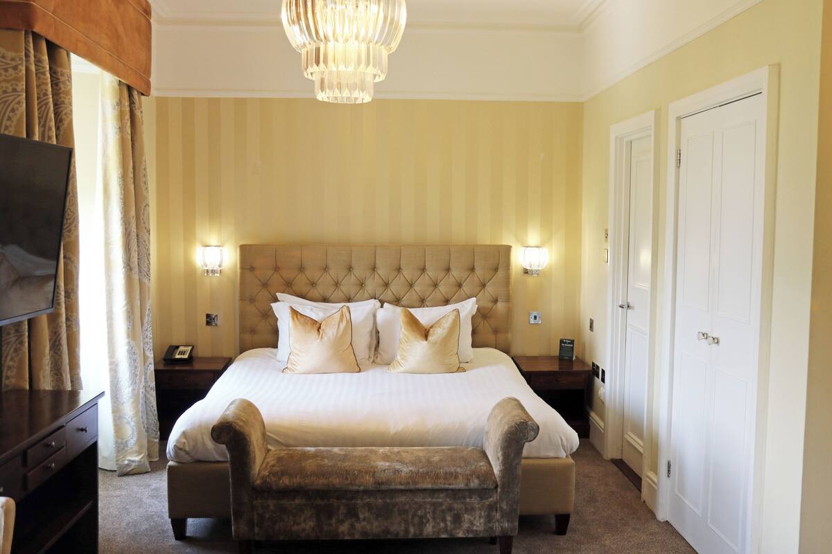 Quarto de hotel com uma cama grande, duas luminárias laterais, uma televisão e um candelabro.