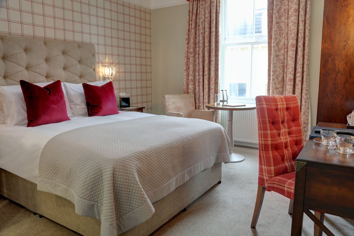 Quarto de hotel com uma cama grande, uma cadeira, uma mesa com copos e uma janela com cortinas.