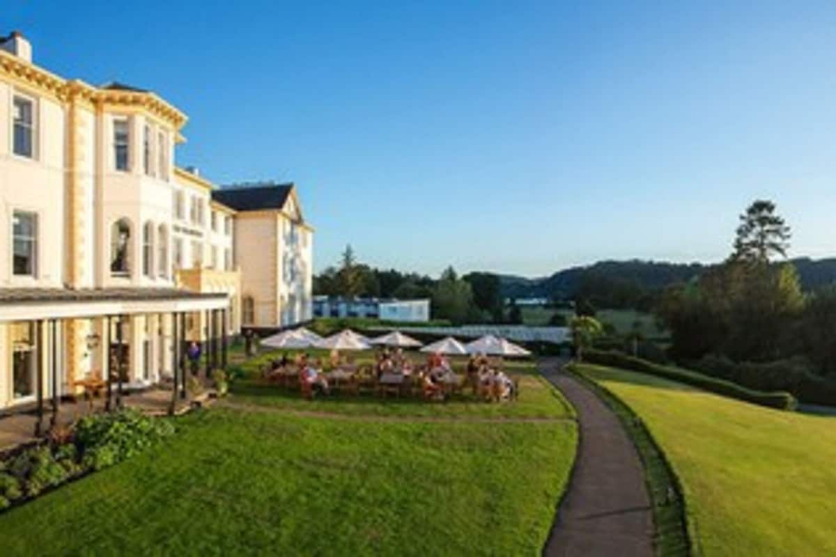 Um edifício grande com gramado, área de estar ao ar livre, caminho e vista para um corpo de água e colinas.