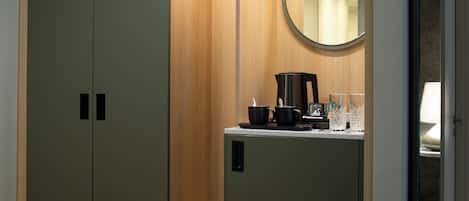 Quarto compacto com parede de painel de madeira, espelho redondo, cozinha compacta com cafeteira e um pequeno armário.