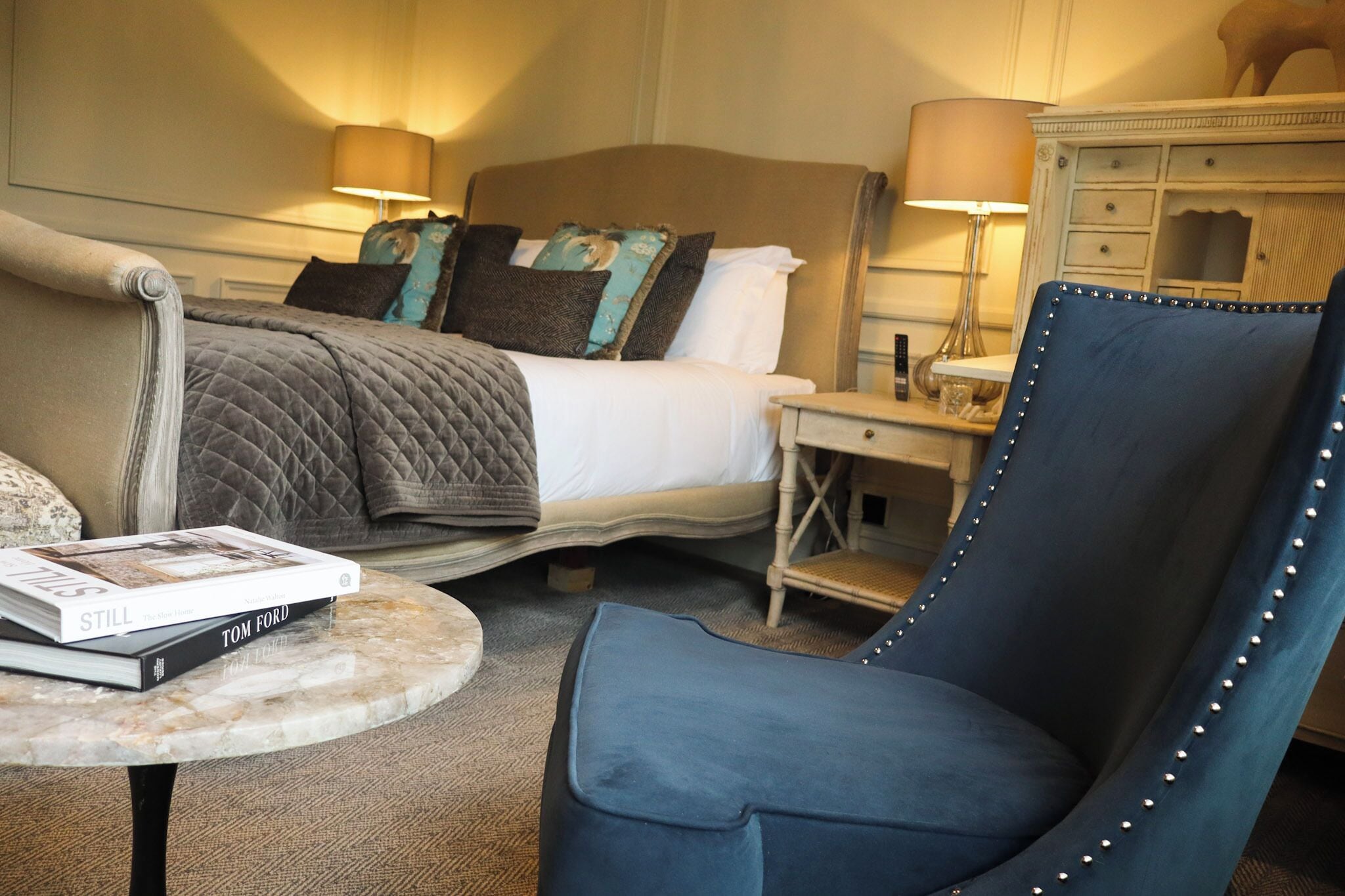Quarto de hotel com cama, cadeira, mesa pequena e cômoda.