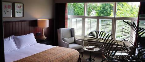Quarto de hotel com cama, poltrona, mesa e janela com vista para o verde.