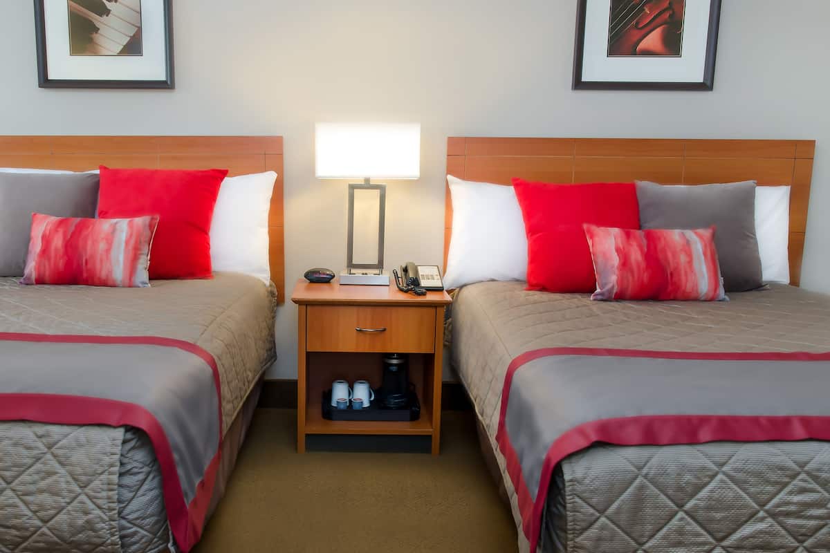 Quarto de hotel com duas camas, cada uma com travesseiro vermelho e branco, uma mesa de cabeceira com abajur e uma gaveta embaixo.