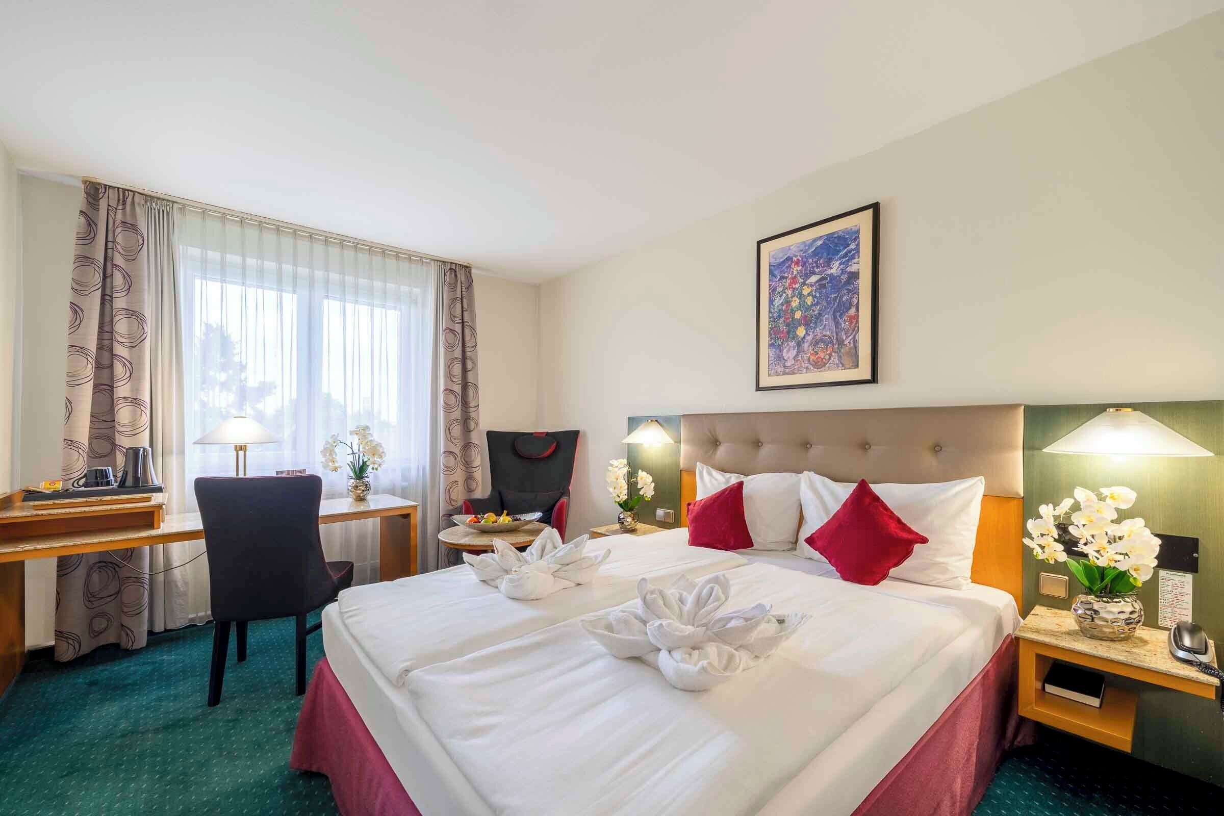 Quarto de hotel com cama, escrivaninha, cadeira e uma pequena mesa com abajur e flores.