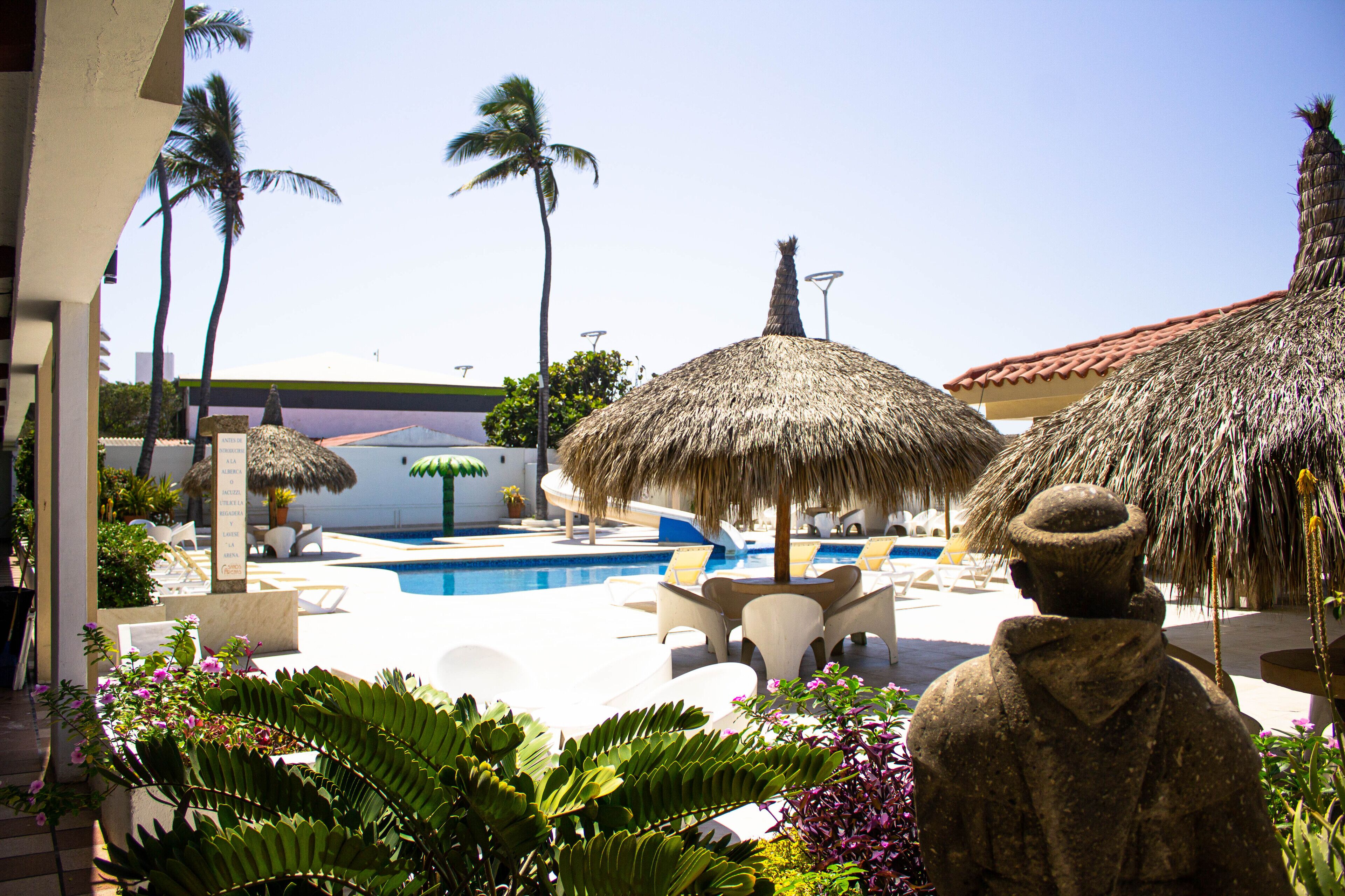 Área de piscina com guarda-sóis de palha, espreguiçadeiras e uma estátua.