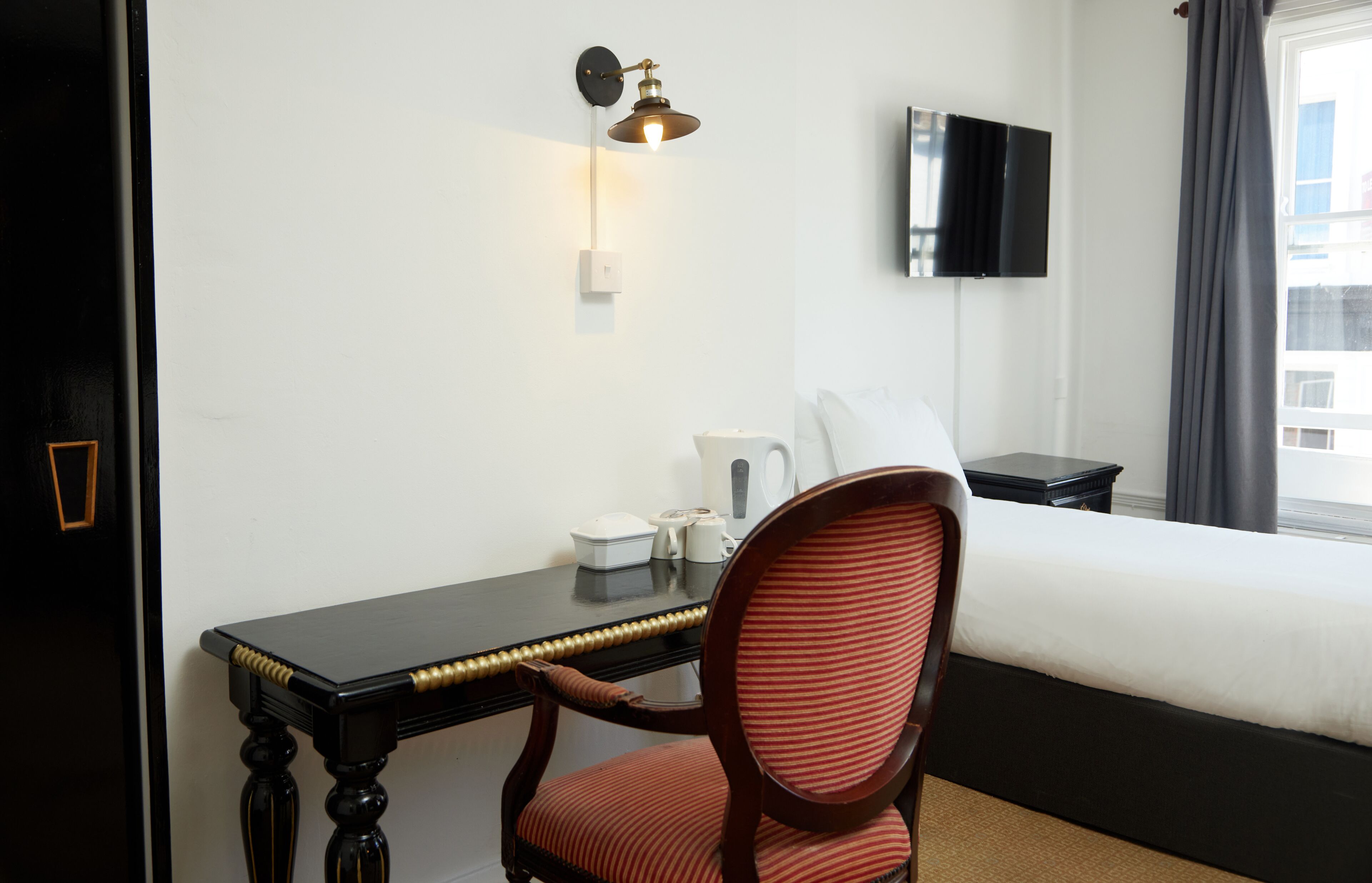 Quarto de hotel com cama, escrivaninha com cadeira, televisão na parede e janela com cortinas.