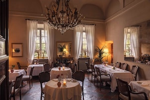 Uma sala de jantar com mesas redondas, cadeiras, lustres e janelas amplas com cortinas.